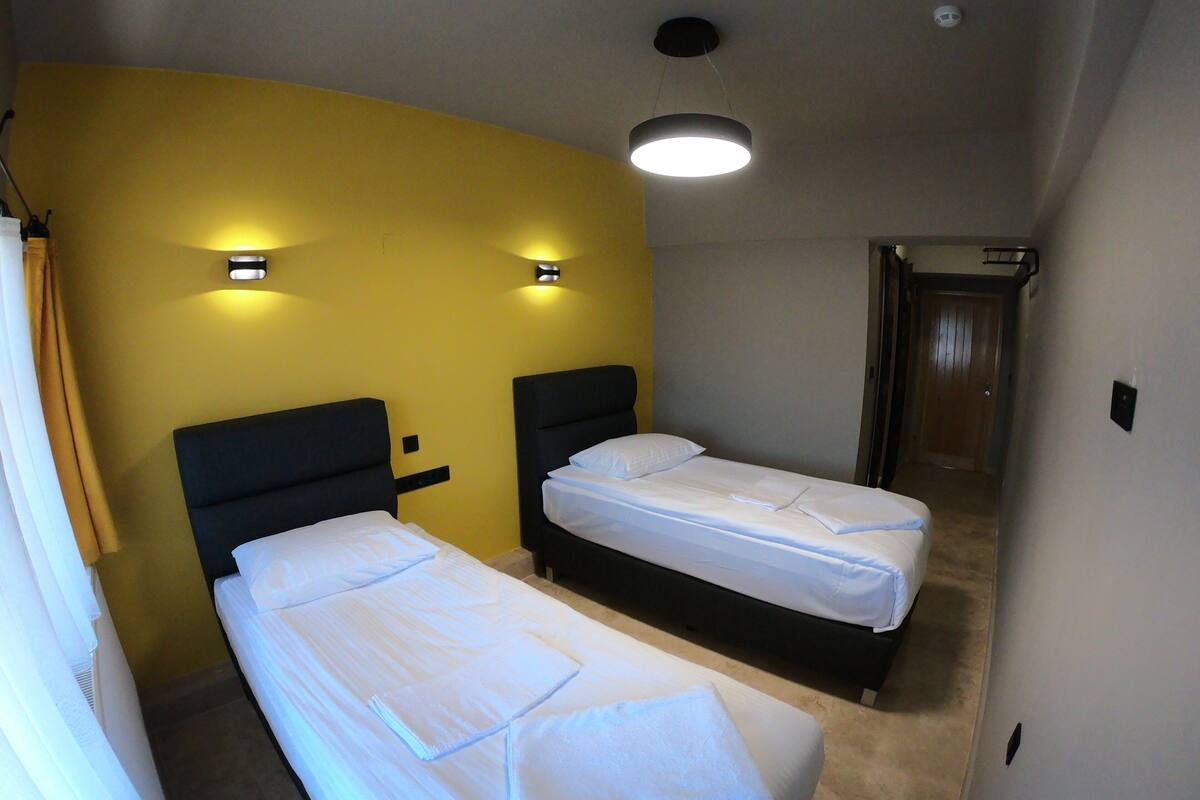 Quarto de hotel com duas camas de solteiro, uma parede de destaque amarela e duas luminárias fixadas na parede.