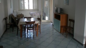 Uma área de refeições com uma mesa e cadeiras, uma televisão sobre um suporte, e uma porta que dá para outro quarto.