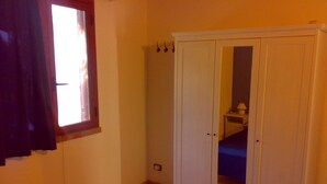 Quarto com guarda-roupa, cama, janela com cortinas e uma pequena mesa com abajur.