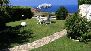 Um jardim com um caminho de pedra, duas cadeiras listradas, uma mesa e um guarda-sol.