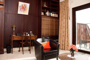 Quarto com escrivaninha de madeira, cadeira de couro preta com almofada laranja, estante e janela com cortinas.