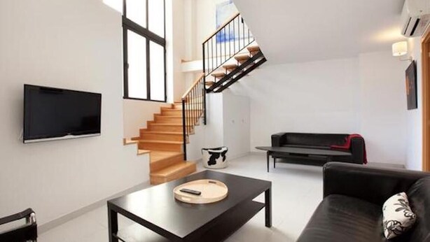 Uma sala de estar moderna com um sofá preto, uma mesa de centro, uma TV fixada na parede e uma escada com degraus de madeira.