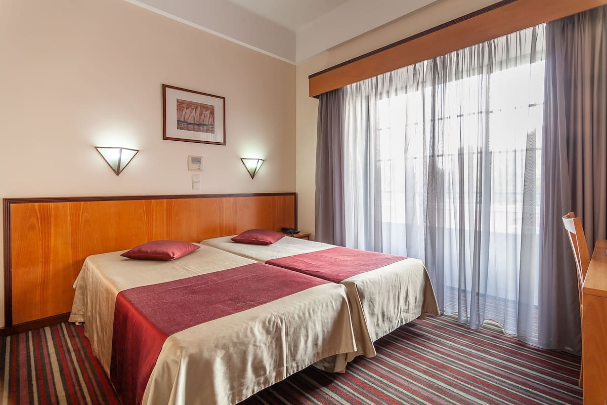 Quarto de hotel com duas camas, cabeceira de madeira, uma janela grande com cortinas leves e um tapete listrado.