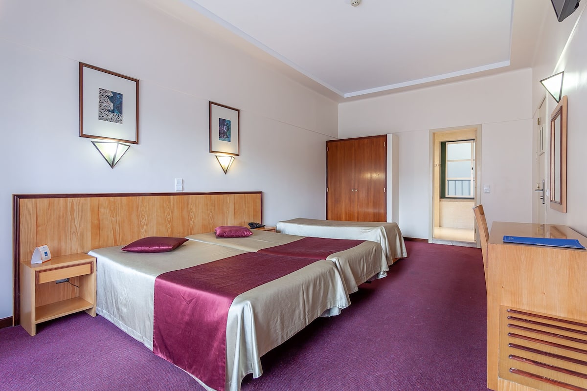 Quarto de hotel com duas camas, uma escrivaninha e um guarda-roupa.