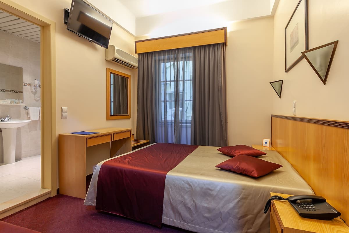 Quarto de hotel com cama, escrivaninha, telefone, espelho e janela com cortinas.