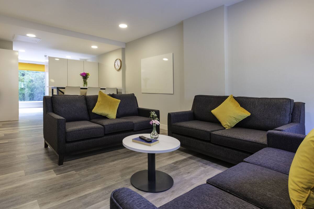 Uma sala de estar moderna com dois sofás cinzentos, cada um com uma almofada amarela, e uma mesa de centro redonda com um vaso e um livro.