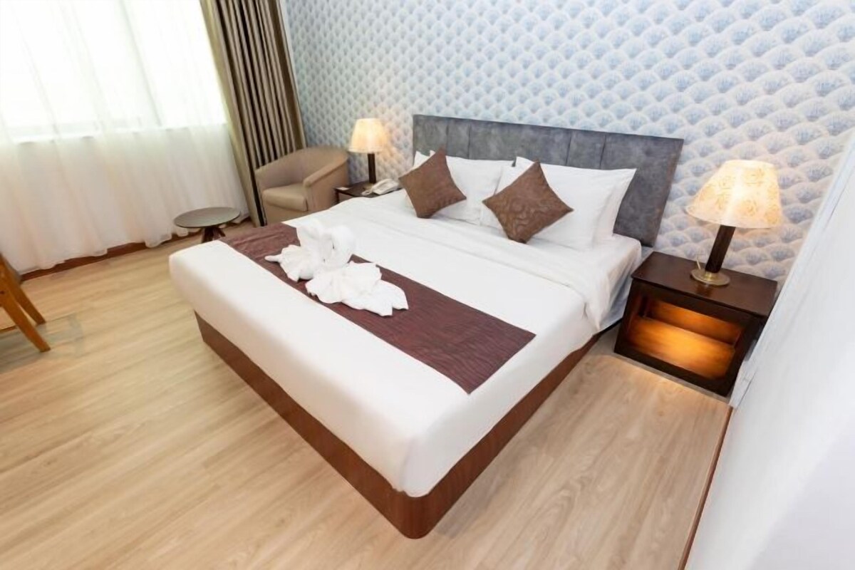 Uma cama bem arrumada com lençóis brancos e almofadas castanhas, uma mesa de cabeceira com abajur e uma cadeira em quarto de hotel com papel de parede estampado e piso de madeira.