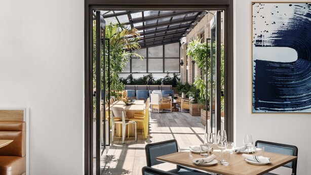 Uma área de jantar moderna com uma mesa de madeira posta para dois, rodeada por cadeiras. Há uma porta de vidro que dá para um pátio ao ar livre com assentos e plantas.