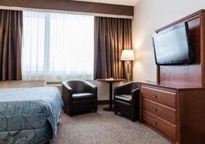 Quarto de hotel com cama, duas poltronas, escrivaninha, luminária e televisão.