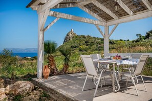 Uma área de estar coberta ao ar livre, com mesa e cadeiras, oferece vista para uma paisagem deslumbrante, incluindo o mar e colinas.