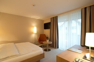 Quarto de hotel com cama, escrivaninha, cadeira, uma pequena mesa com uma planta e televisão na parede.