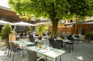 Um café ao ar livre com mesas e cadeiras, cercado por árvores e plantas em vasos.