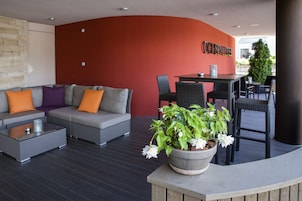 Uma área de estar moderna ao ar livre com um sofá cinza, almofadas laranja, uma mesa de centro de vidro e um vaso com flores brancas.