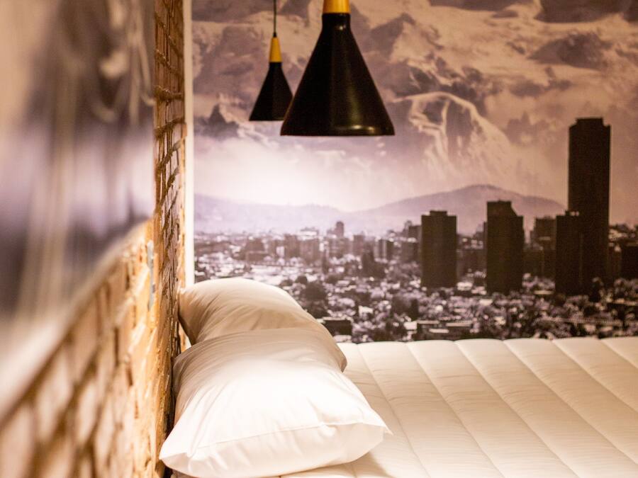 Quarto com um mural da paisagem urbana na parede, uma cama com roupa de cama branca e duas luminárias pendentes pretas.