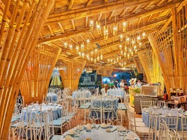 Uma área de refeições interna com estruturas de teto em bambu, cadeiras de metal branco e mesas postas para uma refeição.