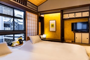 Quarto de hotel com cama, almofadas, mesa de cabeceira com abajur, televisão e porta deslizante com vista para prédios.