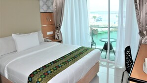 Quarto de hotel com uma cama grande, uma mesa de trabalho e vista para o mar através de uma porta de vidro.