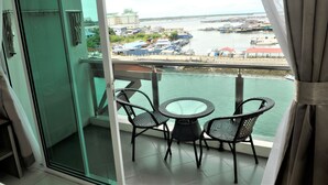 Uma varanda com duas cadeiras de vime e uma mesa pequena, com vista para uma marina com barcos e edifícios.