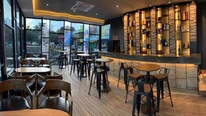 Uma área de bar moderna com mesas e cadeiras de madeira, parede de pedra com prateleiras de garrafas e grandes janelas com vista para a rua.
