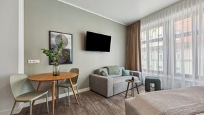 Quarto de hotel moderno com sofá cinzento, uma mesa redonda pequena, televisão na parede e uma janela grande com cortinas leves.