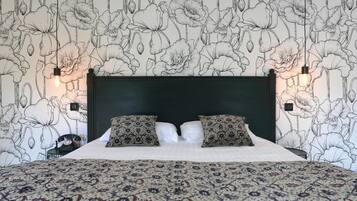 Uma cama com colcha estampada, cabeceira escura e duas almofadas com motivos florais. Há duas luminárias fixadas na parede, uma de cada lado da cama.