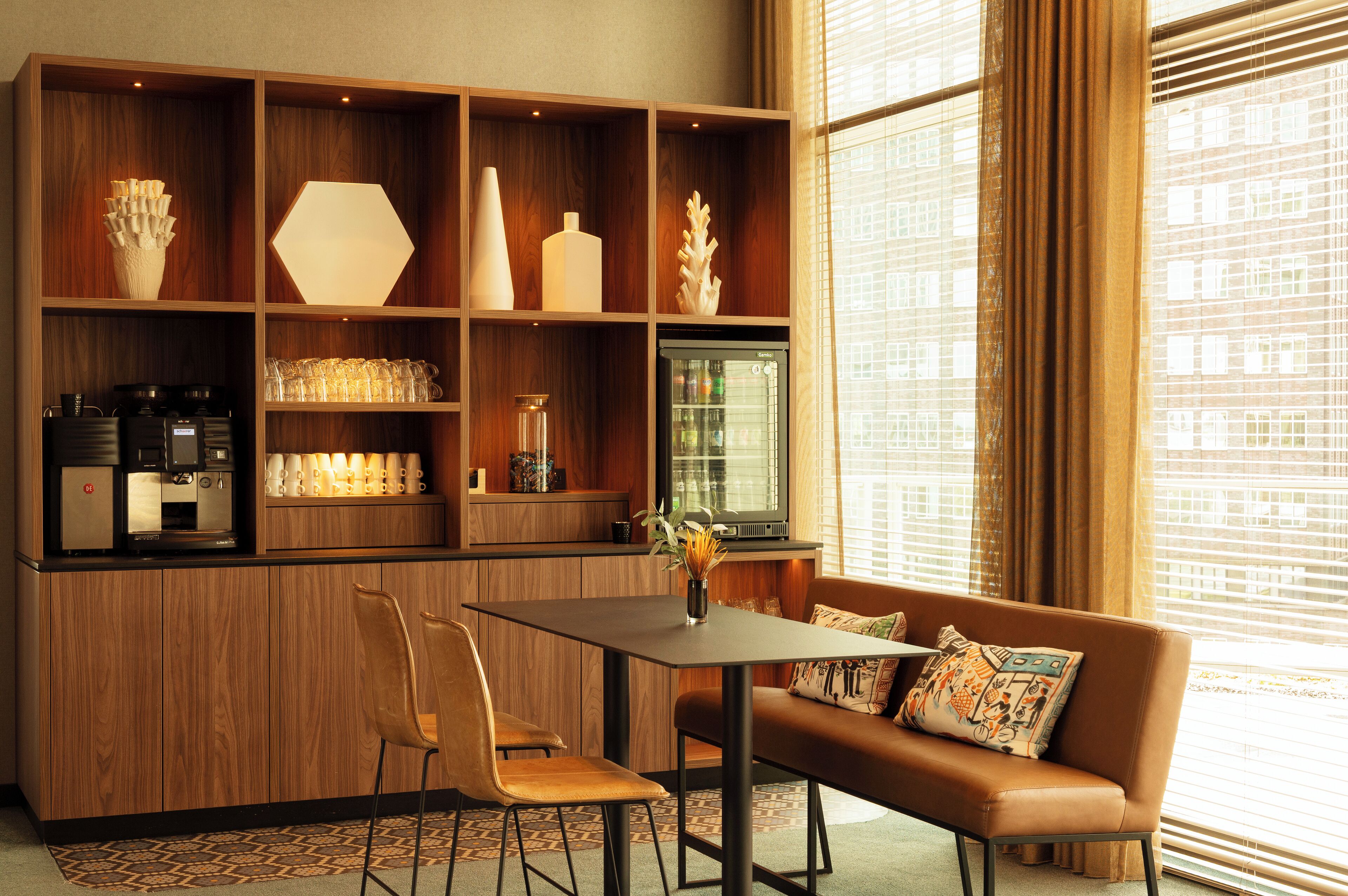 Quarto de hotel moderno com um armário de madeira, uma máquina de café e um sofá com almofadas decorativas.