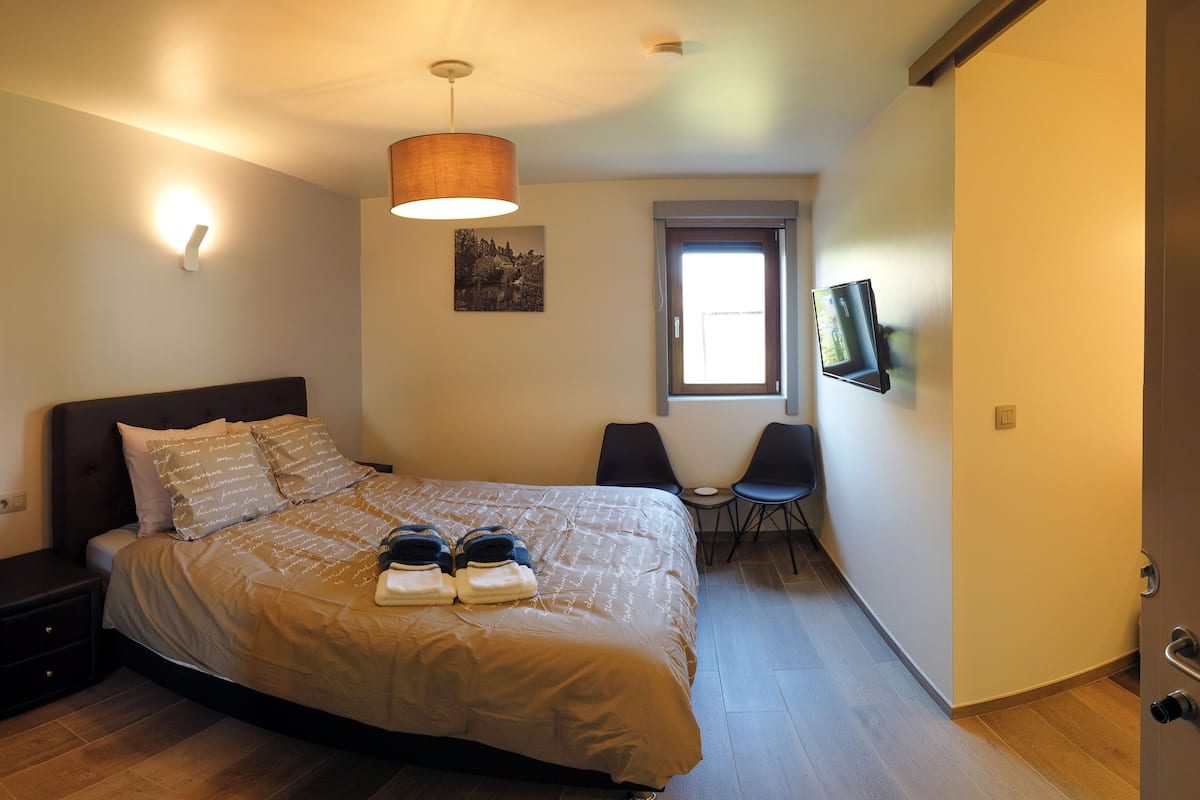 Quarto com cama, escrivaninha, cadeira, televisão e janela.