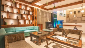 Uma sala de estar ampla com um sofá verde, cadeiras de vime e uma mesa de centro de vidro. Há prateleiras de madeira com livros e objetos decorativos, e uma televisão é visível ao fundo.