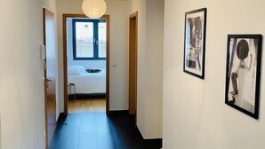 Um corredor com chão em mosaico preto, paredes brancas e uma luminária grande no teto. Há quadros nas paredes e uma cama visível através de uma porta aberta no final.