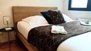 Uma cama bem arrumada com um edredom de padrão geométrico, uma mesinha de cabeceira com abajur e uma janela com luz natural.
