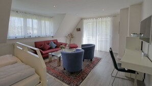 Uma sala de estar moderna com um sofá vermelho, duas poltronas azuis, uma mesa de centro de vidro e uma escrivaninha branca com uma cadeira.