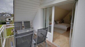 Uma varanda com mesa e cadeiras, porta de vidro deslizante que dá para um quarto com cama e mesa de cabeceira.