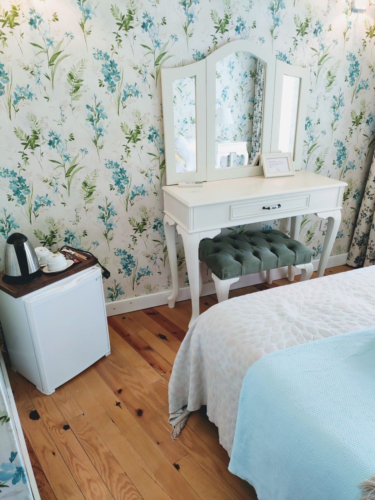 Quarto com penteadeira branca, espelho, um banquinho estofado verde, pequena geladeira branca e cama com manta azul clara.