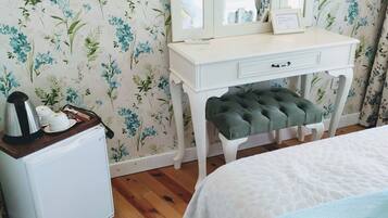 Quarto com penteadeira branca, espelho, um banquinho estofado verde, pequena geladeira branca e cama com manta azul clara.