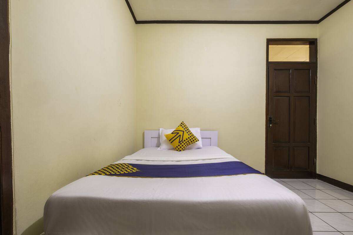 Uma cama de solteiro com colchão branco, edredom azul e amarelo, e travesseiro com padrão geométrico amarelo. Há uma porta de madeira com uma janela acima.