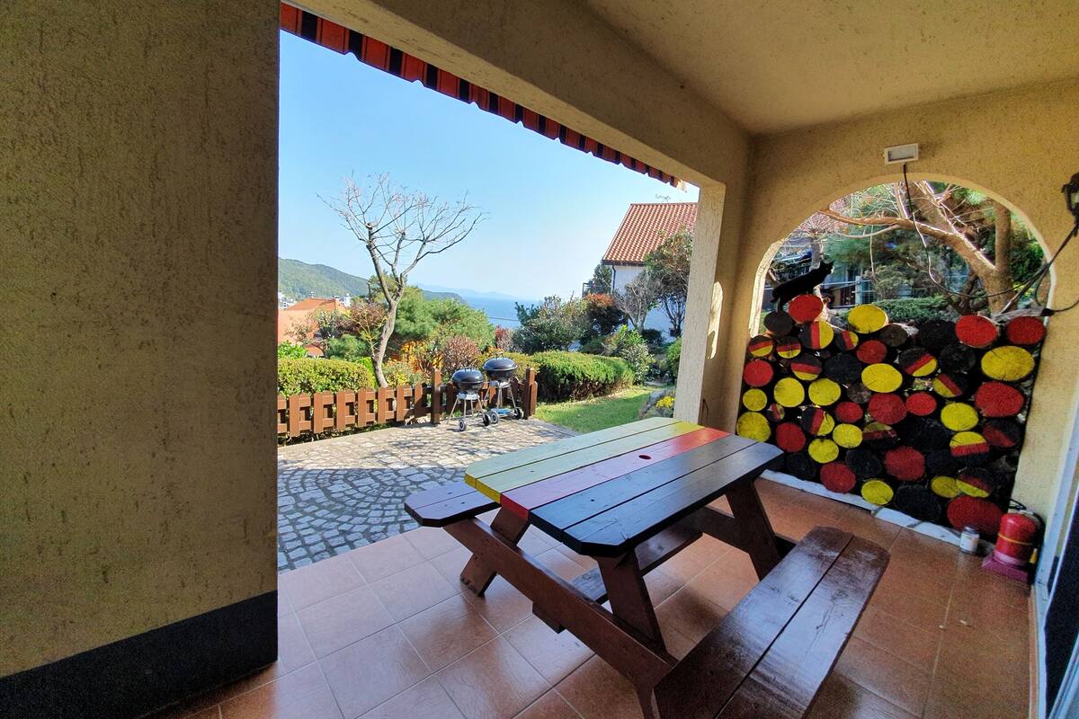Um pátio externo com mesa e bancos de madeira, piso ladrilhado e vista para um jardim e edifícios ao longe.