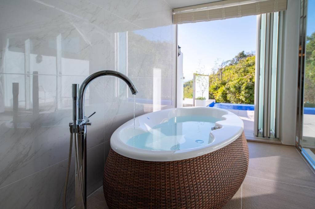 Uma casa de banho moderna com uma banheira independente, cabine de duche com parede de vidro e vista para uma piscina e vegetação.