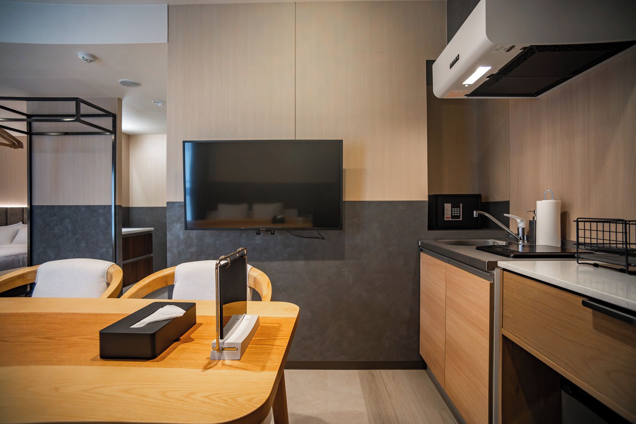 Quarto de hotel moderno com uma mesa de jantar de madeira, uma televisão de tela plana na parede, uma pequena cozinha com pia e cofre, e uma cama com cabeceira.