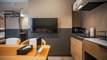 Quarto de hotel moderno com uma mesa de jantar de madeira, uma televisão de tela plana na parede, uma pequena cozinha com pia e cofre, e uma cama com cabeceira.