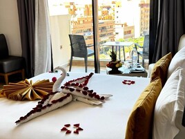 Quarto de hotel com cama, uma cadeira, uma mesa com uma fruteira e uma garrafa, e uma varanda com vista para prédios.
