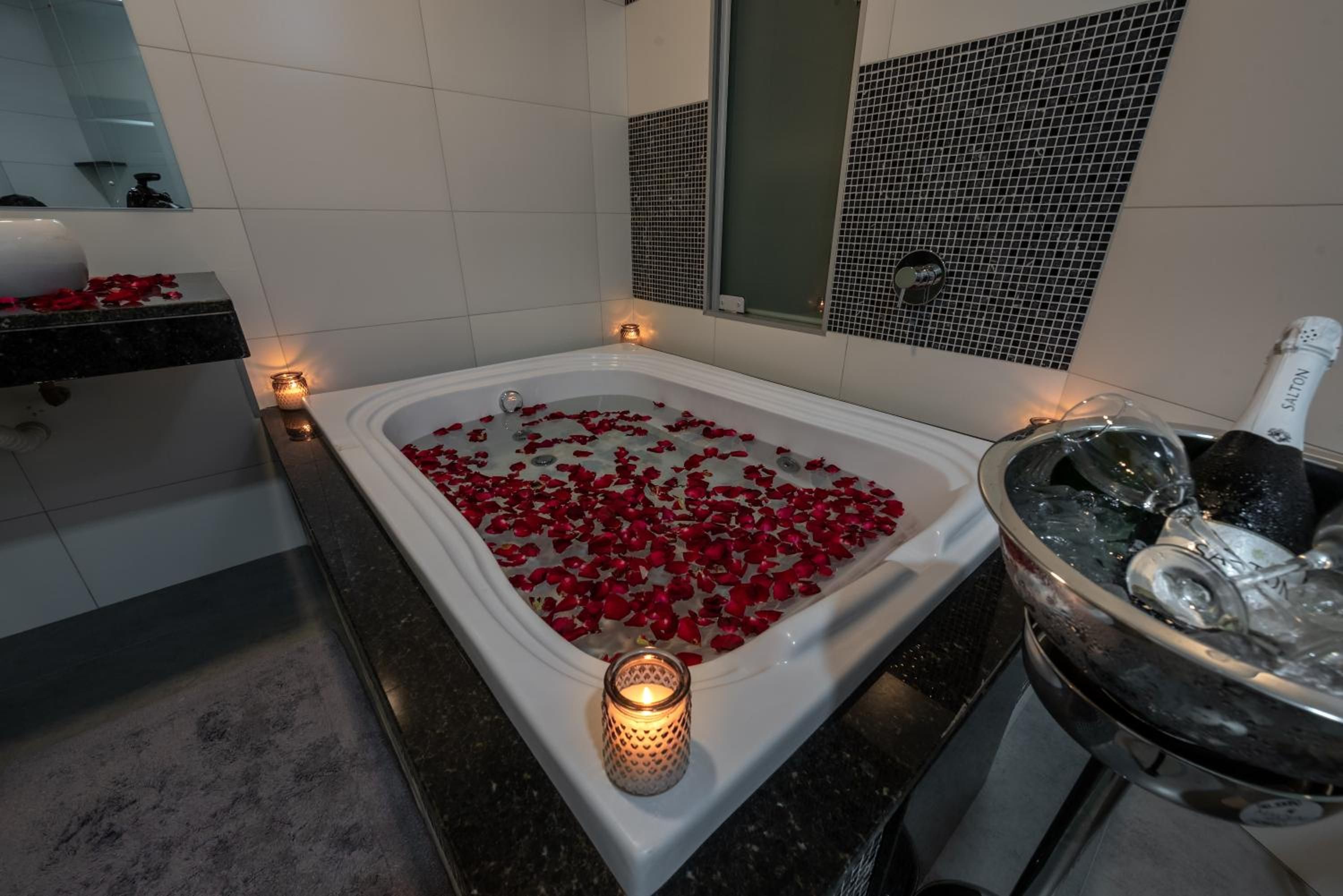 Um banheiro com banheira cheia de pétalas de rosa, uma vela acesa e um espelho sobre a banheira.