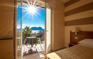 Quarto de hotel com cama, mesa de cabeceira, luminária e varanda com mesa e cadeiras.