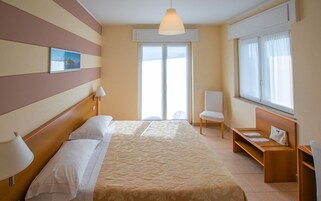Quarto de hotel com cama, luminárias de cabeceira, uma cadeira, escrivaninha e uma janela grande.