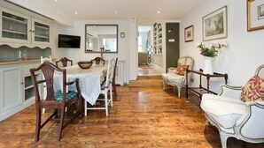 Uma área de refeições com mesa e cadeiras de madeira, uma cozinha com armários e uma televisão, e uma sala de estar com sofá e um vaso de flores.