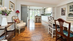 Cozinha espaçosa com armários brancos, área de refeições com mesa e cadeiras de madeira, e um espaço de estar acolhedor com uma cadeira estampada e um vaso de flores.