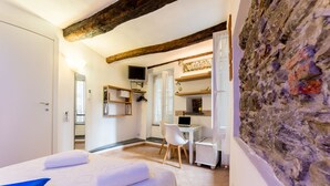 Quarto com cama, escrivaninha e cadeira. Apresenta uma parede de pedra e uma viga de madeira no teto.