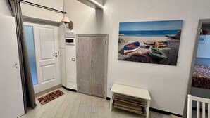 Um corredor com uma porta branca, um banco, um quadro de barcos numa praia e um radiador.