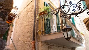 Uma rua estreita com um edifício que apresenta uma varanda com persianas verdes, grades de ferro forjado e um candeeiro suspenso.