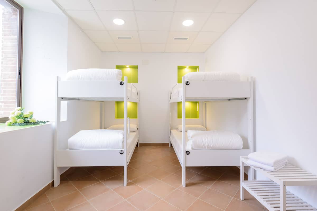 Quarto com duas camas beliche, cada uma com um número lateral, um pequeno banco com uma toalha e uma janela com vista para o verde.