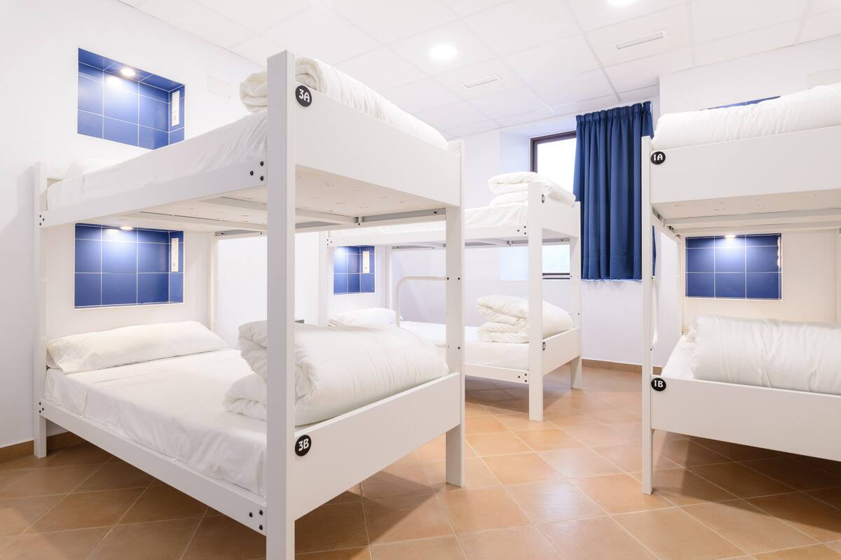 Um dormitório com três beliches, cada um com etiqueta de número. O espaço possui piso de azulejos e uma janela com cortinas azuis.